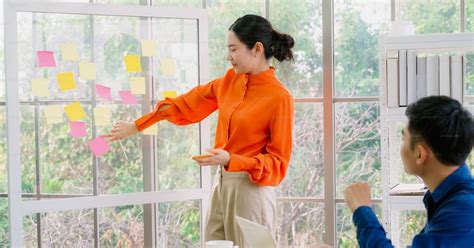 비즈니스 사람들은 사무실의 프로젝트 계획 보드에서 일하고 동료 친구와 대화하여 프로젝트 개발을 분석합니다 그들은 유리 벽에 게시 된 스티커 메모를 사용하여 정리합니다