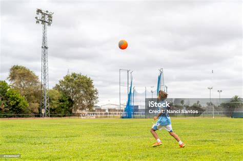 파란색 셔츠와 흰색 반바지를 입은 어린 축구 선수가 밝은 주황색 공을 하늘로 차며 뛰어오르는 모습은 운동장 연습 중 운동 에너지를 보여줍니다 8 9 살에 대한 스톡 사진 및