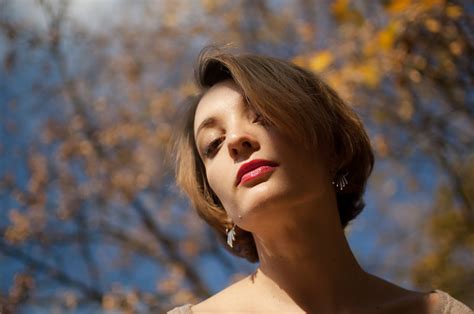 푸른 하늘과 노란 잎 배경이있는 나무를 배경으로 포즈를 취하는 실버 귀걸이와 비게 드레스를 입은 짧은 머리를 가진 아름다운 젊은 아가씨의 야외 초상화가 가을 공원에서 시간을