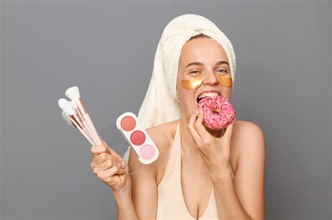 회색 배경 위에 격리된 머리에 상의와 수건을 쓰고 브러시와 아이섀도를 들고 맛있는 도넛을 물고 있는 흥분된 배고픈 웃는 여자의 초상화 30 34세에 대한 스톡 사진 및 기타