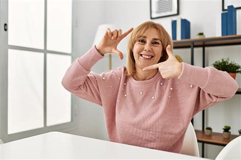 중년 백인 여성은 집에서 테이블에 앉아 앉아 행복한 얼굴로 손과 손가락으로 프레임을 만드는 미소를 지으며 캐주얼 한 옷을 입고 있습니다 창의력과 사진 개념 개념에 대한 스톡