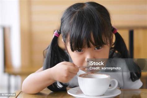 귀여운 아시아 아이 들 또는 아이 여자 들고 숟가락 맛 있는 핫 코코아 또는 가정에서 테이블에 아침에 아침 식사 또는 카페 레스토랑 및 부주의 흰색 컵에 마시는 초콜릿에 대