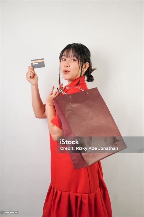 빨간 전통 중국 드레스를 입고 웃는 젊은 아시아 여성이 흰색 배경 위에 고립 된 더미 신용 카드와 종이 쇼핑백을 들고 있습니다