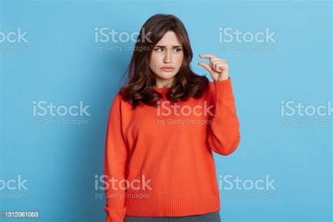 어두운 머리의 여성은 파란색 배경에 고립 된 손가락으로 작은 크기의 제스처를 보여주는 오렌지 스웨터를 입고 불행한 얼굴 표정을 가지고 그녀의 손가락을 본다 20 24세에