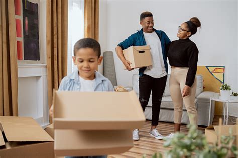 행복한 작은 아프리카 계 미국인 소년은 골판지 상자를 들고 젊은 가족은 구입 한 아파트로 이동 흥분 아들은 개인 소지품을 운반 부모는 서로 자랑스럽게 보고 가득 찬에 대한