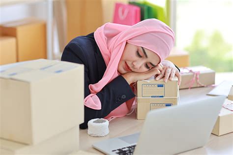 파란 양복과 분홍색 샤프트를 입은 종교적인 아시아 무슬림 여성은 좌절감을 느끼며 테이블에 머리를 얹고 컴퓨터와 소포 상자 배달이 있는 나무 테이블에 앉아 일한다 일하는 동안