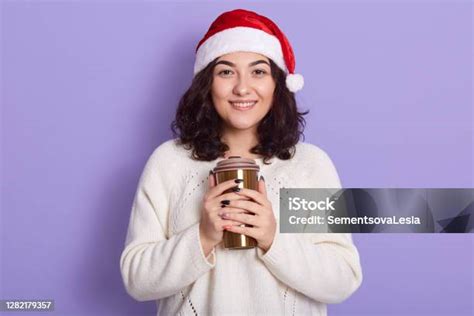 열 잔에서 커피를 마시는 산타 모자와 행복한 젊은 여자 흰색 따뜻한 스웨터드레스 뜨거운 음료를 즐기면서 라일락 배경에 고립 된