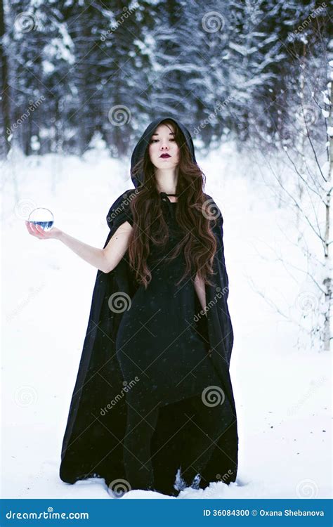 Ведьма или женщина в черном плаще с стеклянным шариком в белом лесе снега Стоковое Фото