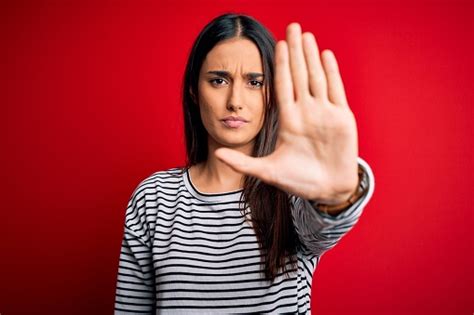 젊은 아름 다운 갈색 머리 여자 캐주얼 줄무늬 티셔츠를 입고 빨간 배경 위에 하 고 중지 손바닥으로 노래 얼굴에 부정적이고 심각한 제스처로 경고 표현 T 셔츠에 대한 스톡