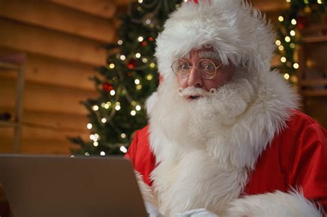 놀란 산타 클로스는 나무 벽과 그의 뒤에 아름다운 크리스마스 트리와 그의 사무실에 앉아있다 그의 노트북에 누군가와 화상 채팅을 갖는 12월에 대한 스톡 사진 및 기타 이미지