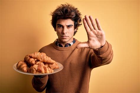 진지하고 자신감있는 표정 방어 제스처로 정지 표지판을하는 열린 손으로 고립 된 노란색 배경 위에 서있는 크루아상과 접시를 들고있는 젊은 잘생긴 남자 경계 표지에 대한 스톡