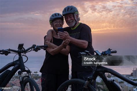 해질녘에 전기 자전거로 언덕에 서있는 사이클링 헬멧을 쓰고 휴대 전화를 보며 웃는 노인 커플 휴가를 즐기는 은퇴자를위한 건강한 활동 2명에 대한 스톡 사진 및 기타 이미지