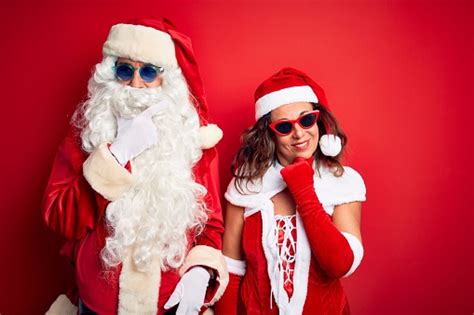 산타 의상과 선글라스를 착용한 중년 부부는 턱에 손을 얹고 미소를 지으며 카메라를 바라보며 고립된 빨간 배경 위에 앉아 있다 긍정적 인 생각 12월에 대한 스톡 사진 및