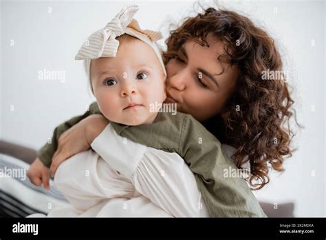 Curly Mother Hugging And Kissing Cheek Of Infant Girl In Headbandstock