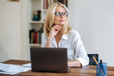 매력적인 성공적인 백인 수석 비즈니스 여성 금발 의여자 흰색 셔츠와 안경을 입고 노트북 앞에 앉아 휴식을 취하고 옆으로 펜을