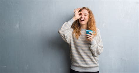 그런 지 회색 벽 손으로 손가락을 통해 찾고 눈에 확인 표시를 하 고 웃 고 행복 한 얼굴로 커피 한 잔을 마시는 동안 젊은 빨강 머리 여자 개념에 대한 스톡 사진 및 기타