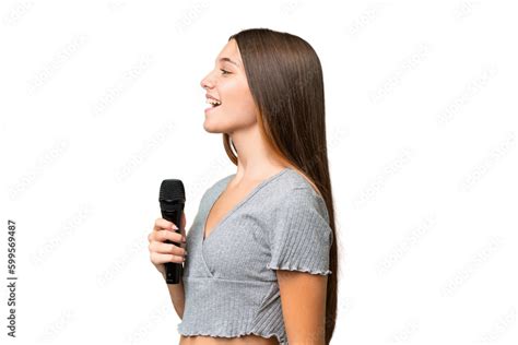 Teenager Singer Girl Picking Up A Microphone Over Isolated Background Laughing In Lateral