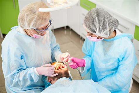 블루 가운에 두 치과 물 주사기의 도움으로는 치과의 남성 고객에 게 치과 치료를 주고 있다 3 명에 대한 스톡 사진 및 기타 이미지 Istock