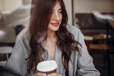 도시 거리에 창을 통해 보이는 카페에 앉아 커피 한잔과 함께 화려한 젊은 여자 아름 다운 머리카락과 미소를 커피와 함께 시간을 즐기고 Hipster 세련 된 소녀 갈색에