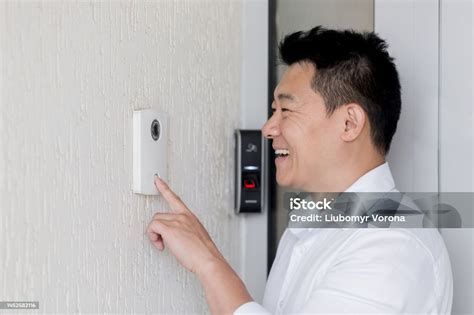젊은 아시아 남자가 손가락으로 집 벽에 전자 초인종을 울립니다 그는 집 문을 엽니다 초인종에 대한 스톡 사진 및 기타 이미지