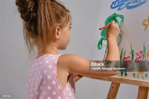 분홍색 드레스를 입은 어린 소녀 아티스트가 이젤 뒤에 서서 흰색 스튜디오 배경에 고립 된 캔버스에 브러시로 그림을 그리고