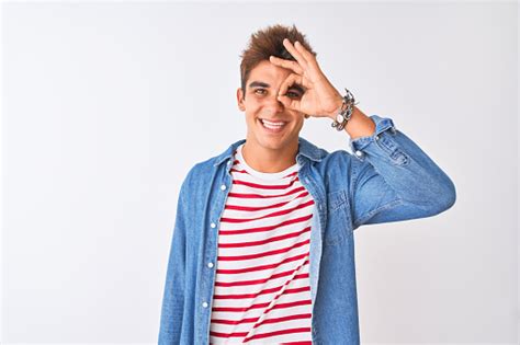 줄무늬 티셔츠와 데님 셔츠를 입은 젊은 잘생긴 남자는 고립 된 흰색 배경 위에 손을 미소 짓고 행복한 얼굴로 손가락을 통해 눈을 보고 확인 제스처를하고 있습니다 T 셔츠에