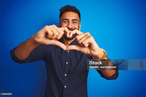 고립 된 파란색 배경 위에 서있는 셔츠를 입은 젊은 인도의 우아한 남자는 손으로 심장 기호와 모양을 보여주는 사랑에 미소 로맨틱 한 개념 경영자에 대한 스톡 사진 및 기타