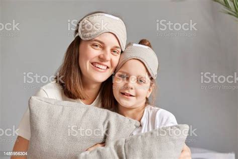 귀여운 딸과 함께 침대에 앉아 있는 쾌활한 어머니는 베개를 들고 미소를 지으며 카메라를 바라보며 자유 시간을 함께 즐기고 있습니다 2명에 대한 스톡 사진 및 기타 이미지