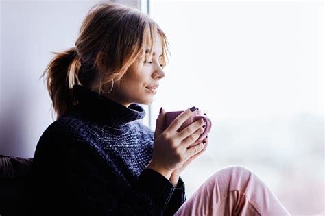 파란 스웨터를 입은 사려 깊은 소녀가 커피 한 잔을 들고 창문 에 앉아 거리를 바라봅니다 20 24세에 대한 스톡 사진 및 기타 이미지 20 24세 가정 생활 가정의