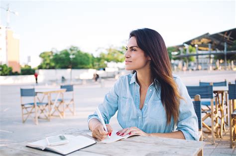 캐주얼 한 옷을 입고 아름다운 사려 깊은 여성 기사를 만들기위한 카피 북에서 메모를 만드는 젊은 30 대 여성은 카페 테라스에서 야외에서 앉아 작업을 설계하기위한 스케치를