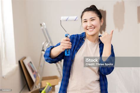 아시아의 아름다움을 지닌 25 세의 소녀는 한 손으로 페인트 롤러를 매우 즐겁게 들고 다른 한 손으로 슈퍼를 보여줍니다 그녀는 꿈의 개조가 그녀의 집에서 시작되었다는 것을