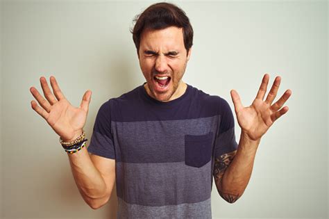 고립 된 흰색 배경 위에 캐주얼 티셔츠를 입고 문신을 한 젊은 잘생긴 남자는 팔을 들고 눈을 감고 흥분을 일으키며 성공을 축하하기 위해 미친 듯이 축하합니다 우승자 컨셉 T