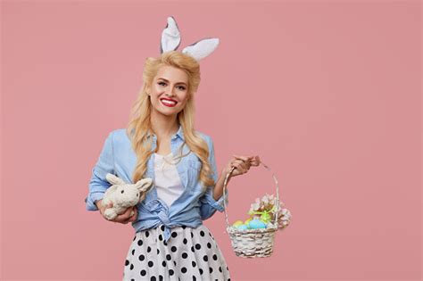행복한 부활절 토끼 귀를 가진 젊은 핀 업 여자는 분홍색 배경에 유기농 색깔의 계란으로 가득 찬 바구니를 들고 있습니다 행복한