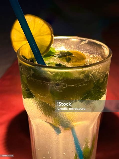 빨간 식탁보 민트 잎 레몬 슬라이스 및 플라스틱 마시는 빨대 결로 거품 밤의 야외 해변 레스토랑에 얼음 조각이 있는 모히토 칵테일의 백라이트 잔의 클로즈업 이미지 전경에