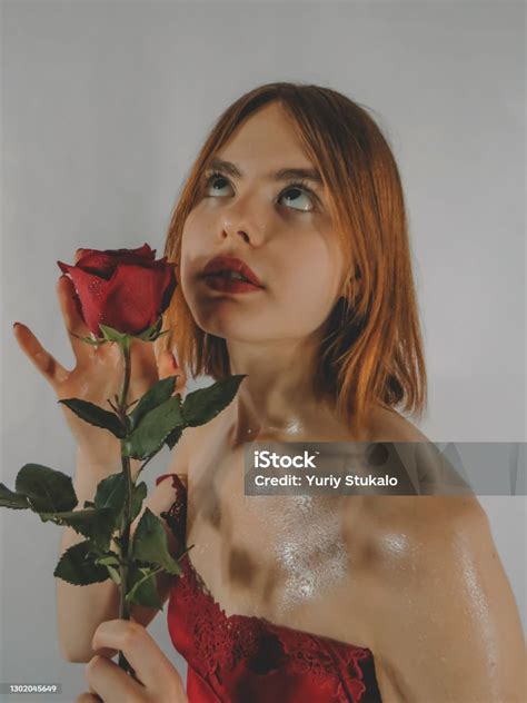 꽃잎에 물 방울이 있는 밝은 빨간 장미의 큰 꽃과 함께 열린 어깨가 있는 버건디 실크 드레스를 입은 매력적인 젊은 빨간 머리의 여성 18 19세에 대한 스톡 사진 및 기타