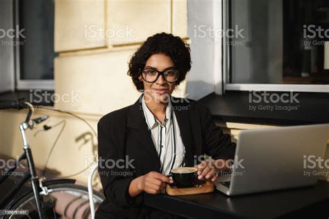 안경을 쓴 아프리카계 미국인 소녀가 노트북과 커피 한 잔을 들고 카페 테이블에 앉아 있다 검은 색 재킷에 어두운 곱슬 머리를