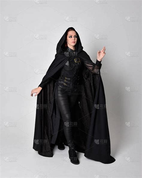 一个红头发的女人穿着黑皮刺客服装和黑色长斗篷的幻想画像。全长站立姿势，与工作室背景隔离