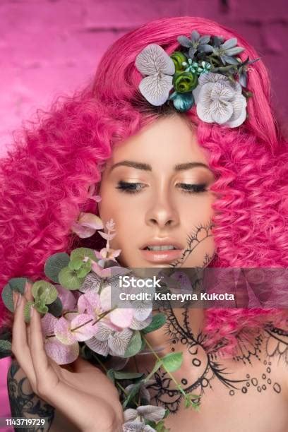 아프리카 스타일 곱슬 밝은 분홍색 머리 문신 얼굴과 목과 꽃 그녀의 머리에 짠 젊은 매력적인 백인 여자 모델입니다 분홍색 배경에 스튜디오에서 사진 감정에 대한 스톡 사진 및