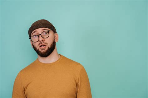 모자를 쓰고 안경을 쓴 스웨터를 입은 낙담한 수염 난 청년이 옆으로 옮겨졌고 얼어붙은 얼굴 표정은 빈 공간이 있는 청록색 스튜디오 배경 위에 서 있습니다 모형 충격을받은