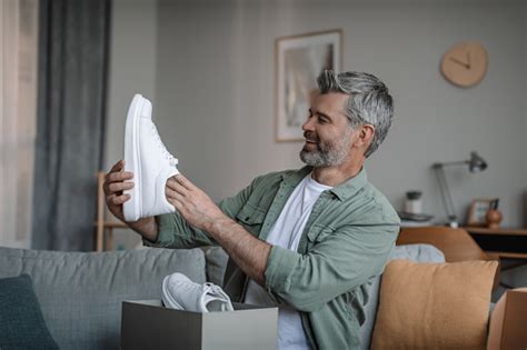 수염을 기른 쾌활한 은퇴한 유럽 남자가 거실 인테리어에 운동화가 있는 소포 상자를 엽니다 스포츠 신발에 대한 스톡 사진 및 기타 이미지 스포츠 신발 구매 새로운