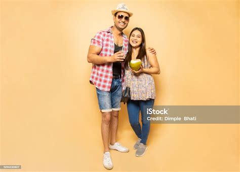 모자와 선글라스를 쓴 쾌활한 젊은 인도 부부는 베이지색 스튜디오 배경에 고립된 코코넛 물과 탄산음료를 마신다 휴일과 휴가 여름 시즌 컨셉 전체 길이 복사 공간 여름에 대한