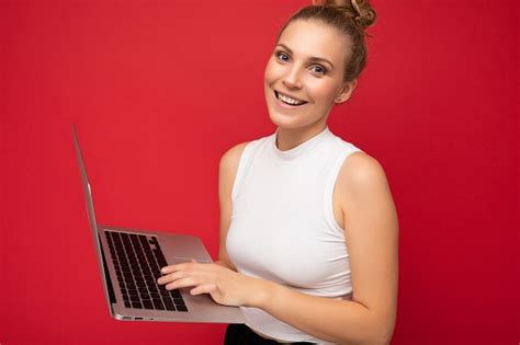 빨간 벽 배경 위에 고립 된 카메라를보고 키보드에 입력 컴퓨터 노트북을 들고 흰색 티셔츠를 입고 모은 머리와 아름다운 미소 금발 젊은 여성의 사이드 프로필 사진 T 셔츠에