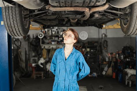 자동차 작업장과 안경을 쓴 여성 정비사가 차고의 차 아래에 서 있습니다 자물쇠 장인에 의한 자동차 검사 파란색 바지에 소녀 복사 공간 검사 보기에 대한 스톡 사진 및 기타