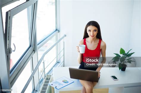사업입니다 아름 다운 젊은 여 자가 옷을 입고 빨간 T셔츠와 블랙 스커트는 컴퓨터에서 일하고 있다 손에 컵입니다 여자는 미소 하 고 테이블에 앉는 다 높은 해상도 T 셔츠에