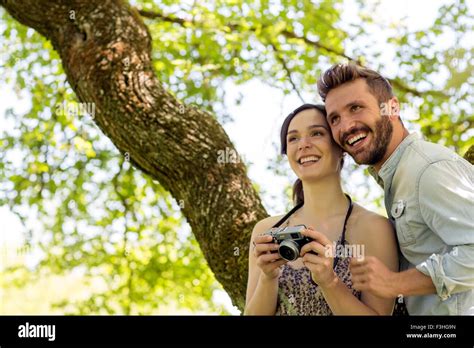 Young Couple Underneath Tree Holding Camera Looking Away Smiling Stock