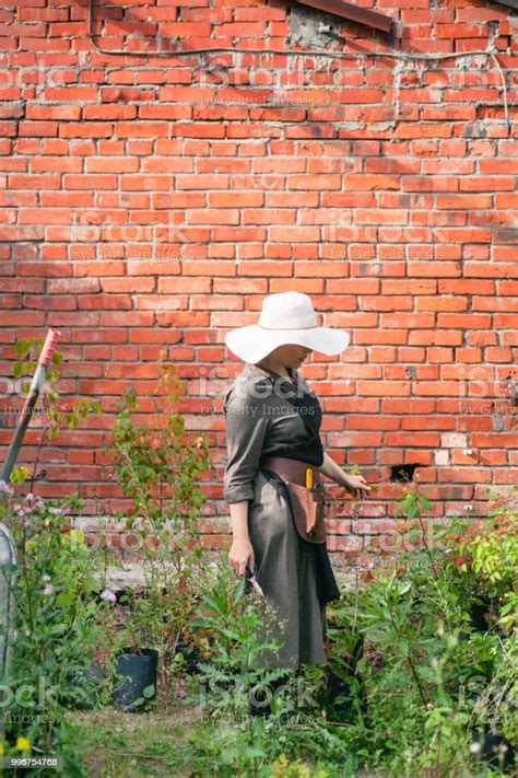 젊은 귀여운 여자를 입고 밀 짚 모자 리넨 드레스 검은 앞치마와 가죽 벨트 플로리스트 도구 정원에서 일하고 도시 원 예 개념