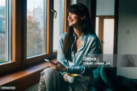 스마트폰을 사용하고 창가에서 커피를 마시는 웃는 여성 사업가 25 29세에 대한 스톡 사진 및 기타 이미지 25 29세 Brand Name Smart Phone 가정