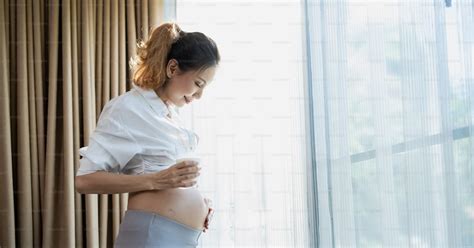 아시아의 아름다운 임신 여자는 건강 관리를 위해 우유 한 잔을 마신다 매력적인 임신 한 소녀는 거실에서 실내에 서서 집에서 행복과 사랑으로 자궁에서 아기를 만지고