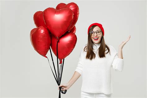 흥분 한 젊은 갈색 머리 여자 스웨터 빨간 모자 안경 생일 휴가 파티를 축하 하는 손을 확산 하 고 흰색 배경 스튜디오 초상화에 고립 된 무리 심장 공기 팽창 헬륨 풍선을