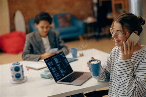 Female Programmer Is Talking Phone While Working On Laptop And Drinking Coffee Sitting At